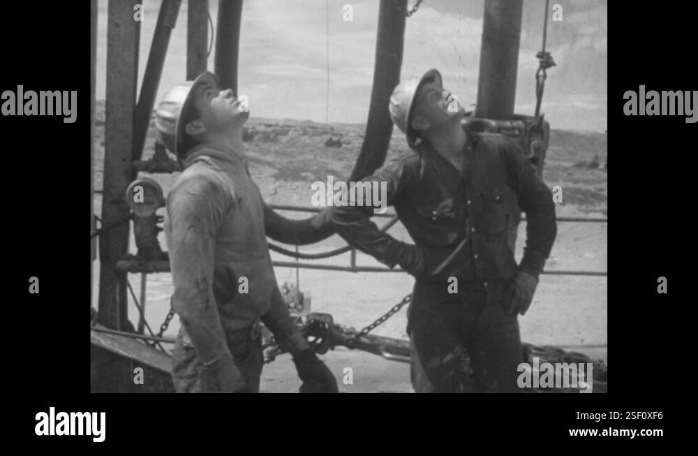 1950s: Man rides on drill bit on pulley up oil derrick. Men and boy on ...