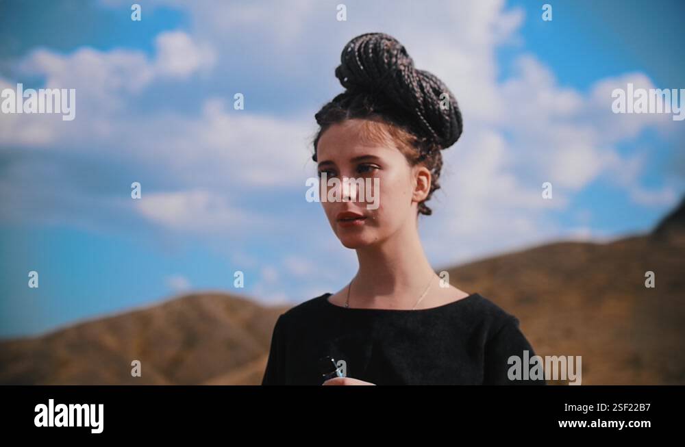 Young beautiful woman with dreadlocks standing on mountain and vaping ...