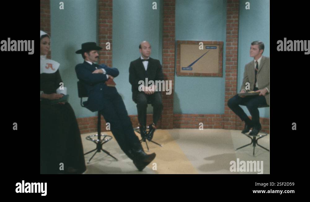 1970s: Film slate. Man in suit sits in chair on stage with three actors ...