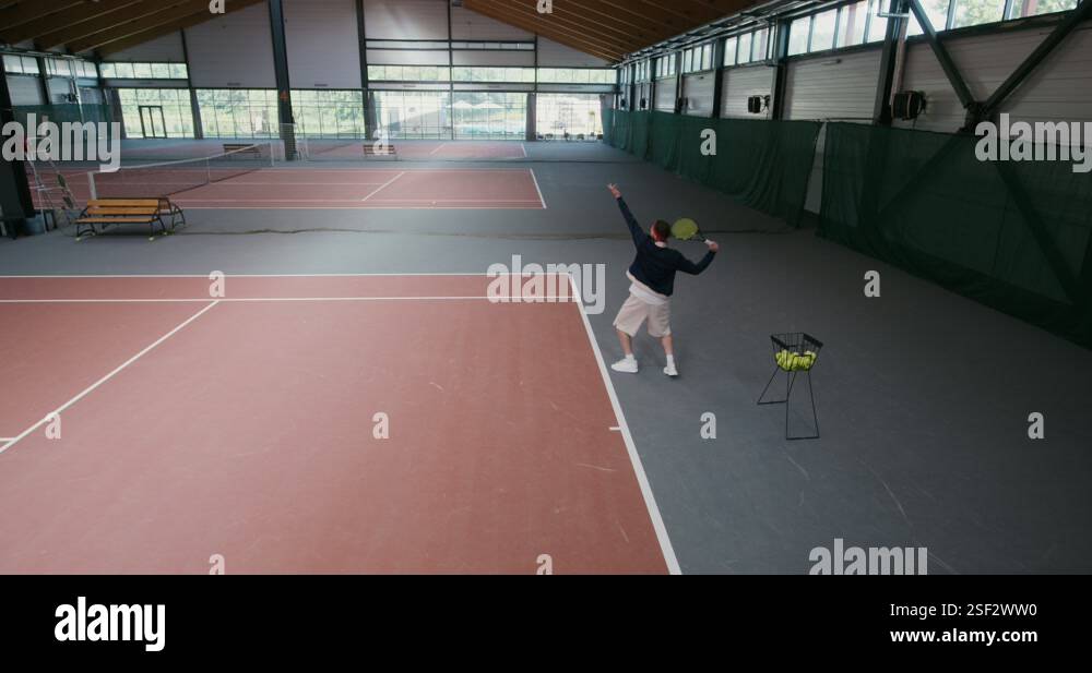 A man plays tennis alone, taking a ball from a basket and tossing it ...