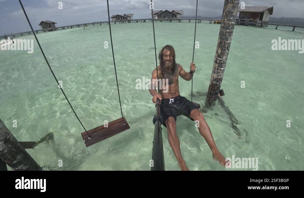 A young, fit and strong man is sitting alone on a swing, smiling with ...
