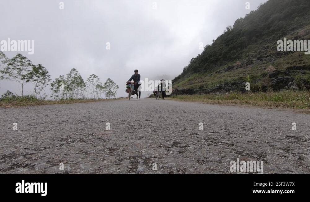 Two cyclist walking towards camera and dragging their cycles loaded ...