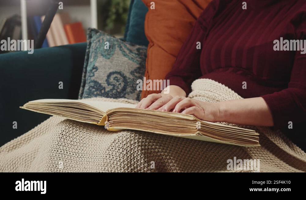 Blind woman reading braille book using fingers, sitting on sofa in ...