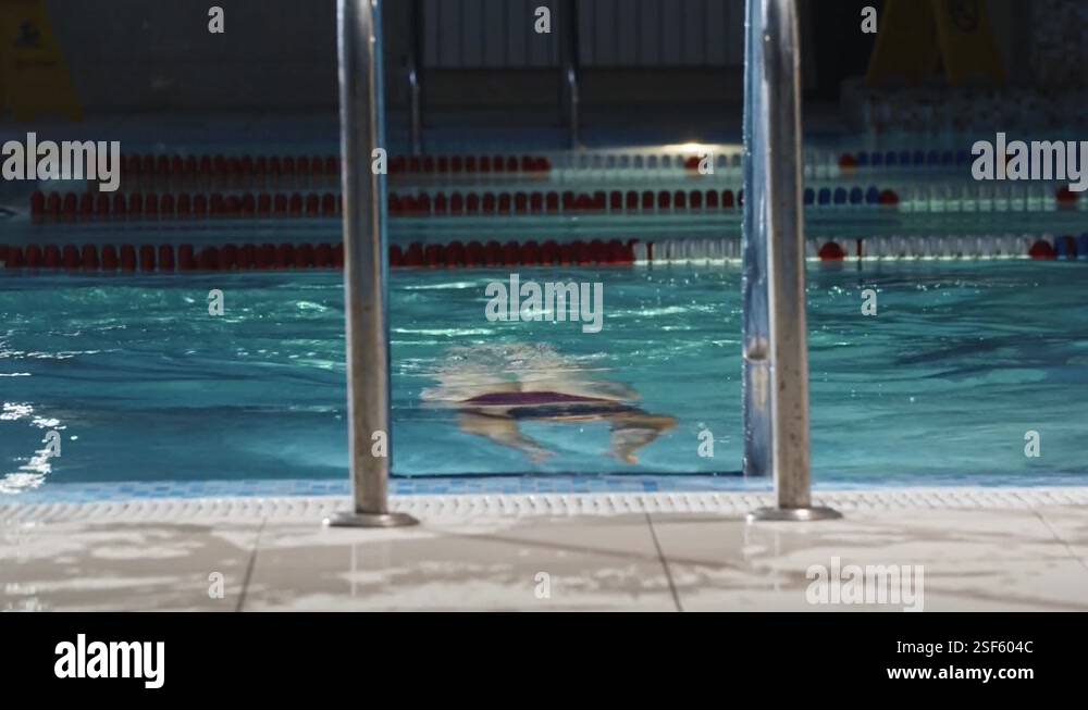 Young woman swimmer in red swimsuit swims to the ladder of a pool and ...