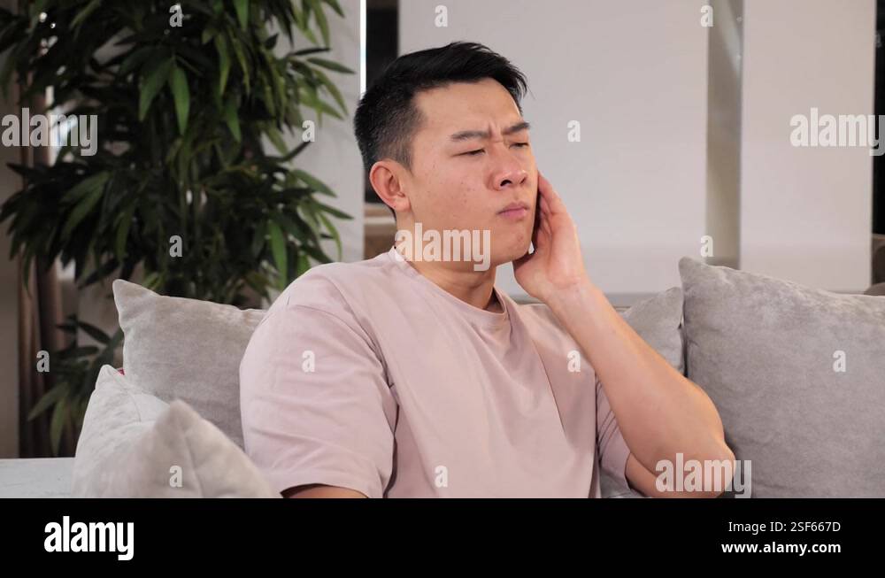 Asian man sitting on sofa uses phone and has sharp toothache dental ...