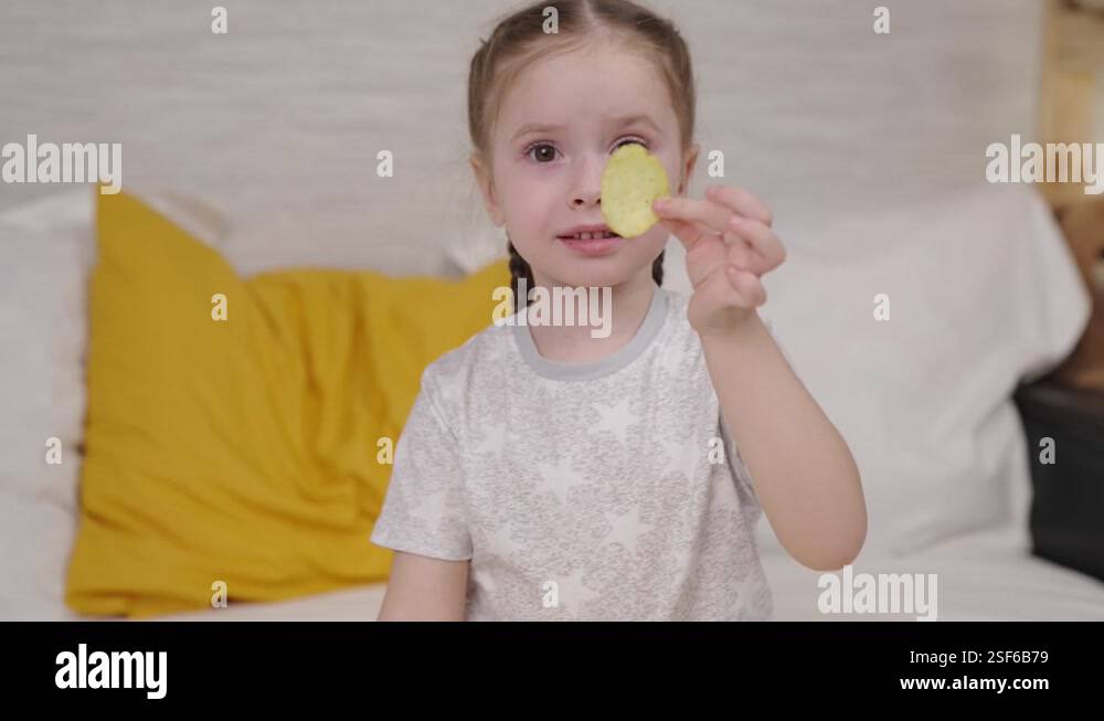 little girl eating potato chips while sitting on the bed, enjoying ...