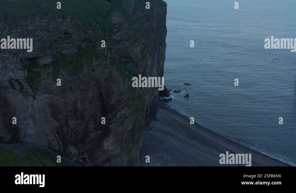 Fly around massive rock at sea coast. Mild waves washing black volcanic ...