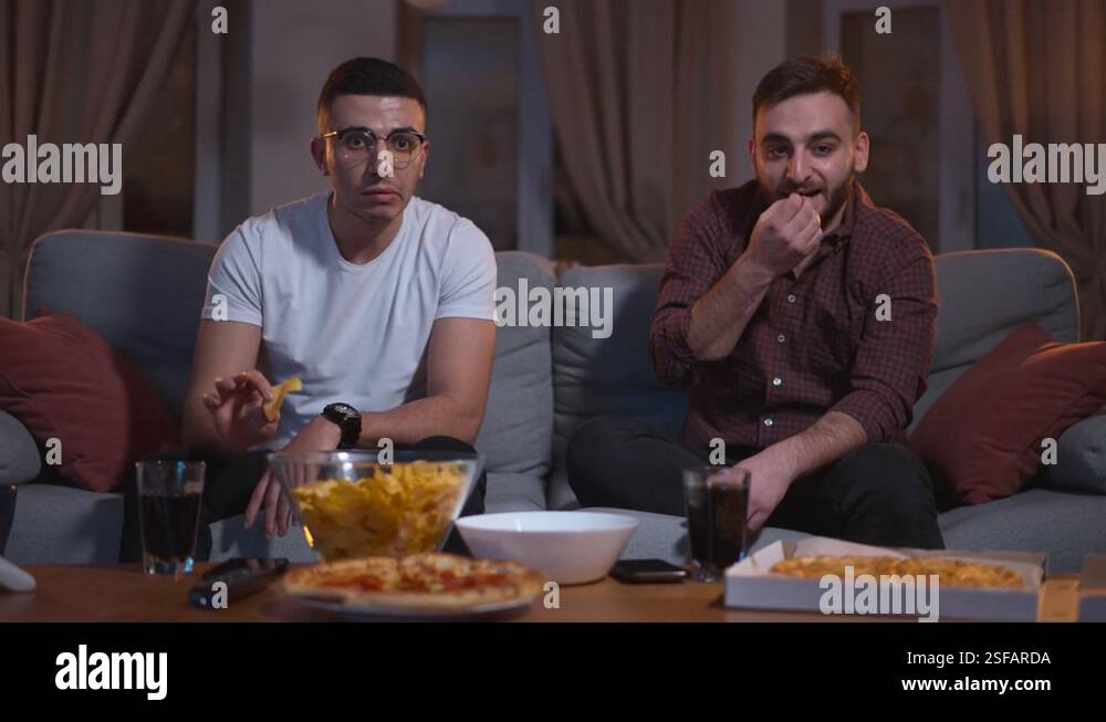 Two young men watching football and eating food while sitting on sofa ...