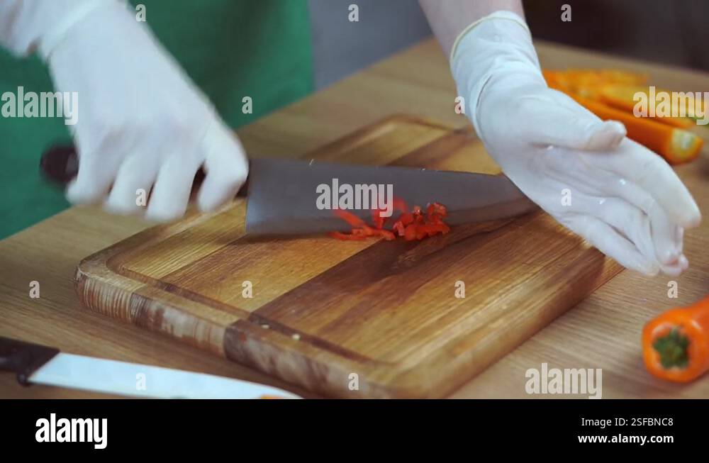 Gloved hands finely chop red chili peppers on a cutting board. Cook ...