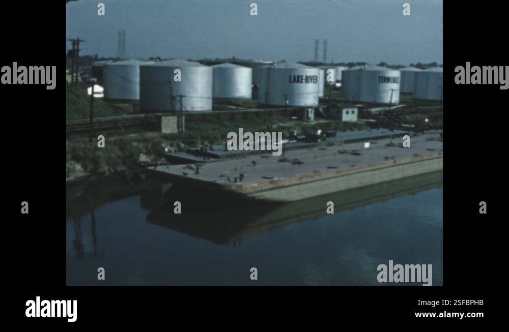 1950s: Large silos on bank of river. Flat docks float on river. Smoke ...
