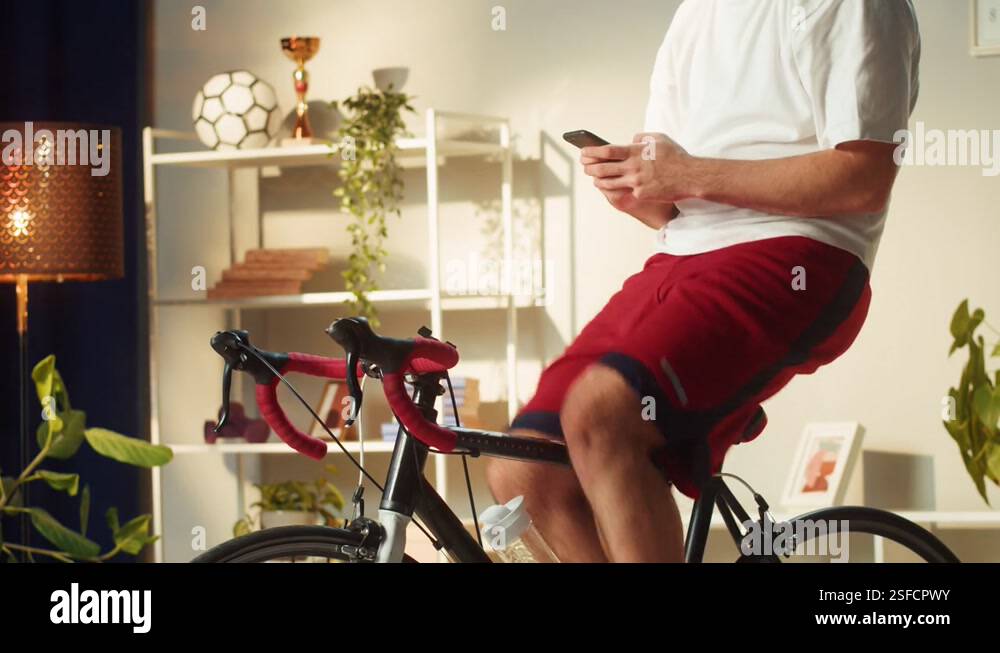 Sportsman riding bicycle and using phone, home gym. Young man cycling ...
