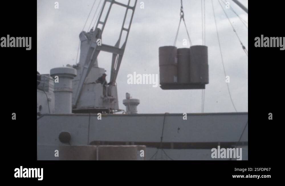 1960s: Man operates crane, lifts barrels onto boat. Man climbs on top ...