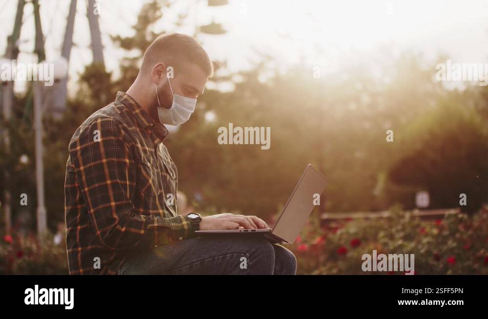 Masked man is typing on laptop while sitting park bench, turns his head ...