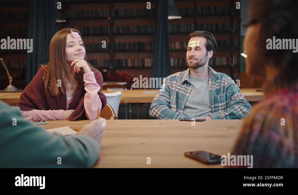 Young people students sitting around table playing funny guessing game ...