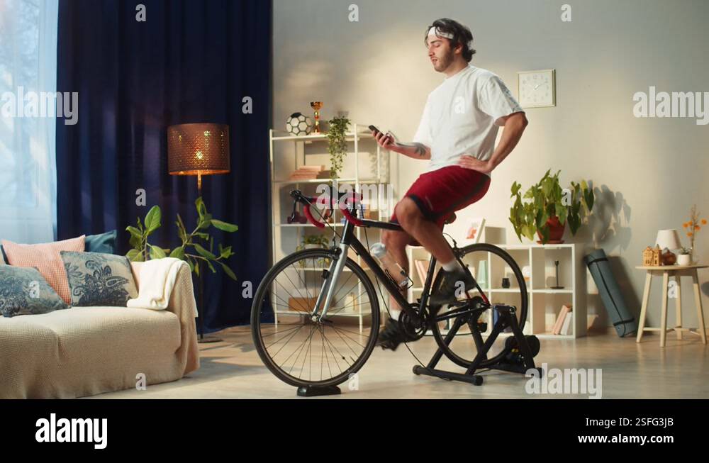Sportsman riding bicycle and using phone, home gym. Young man cycling ...
