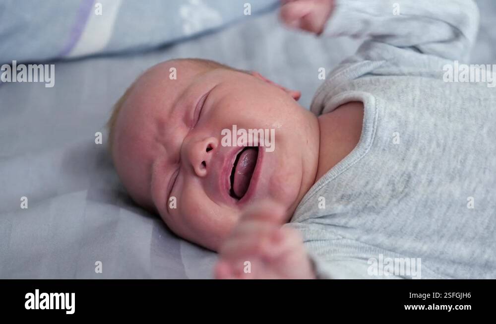 Scared newborn girl in grey bodysuit lies on bed and screams out loudly ...