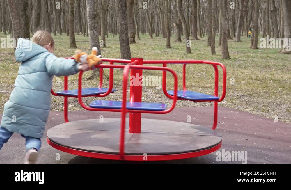 A little girl in a blue coat with a plush toy spins on carousel on ...