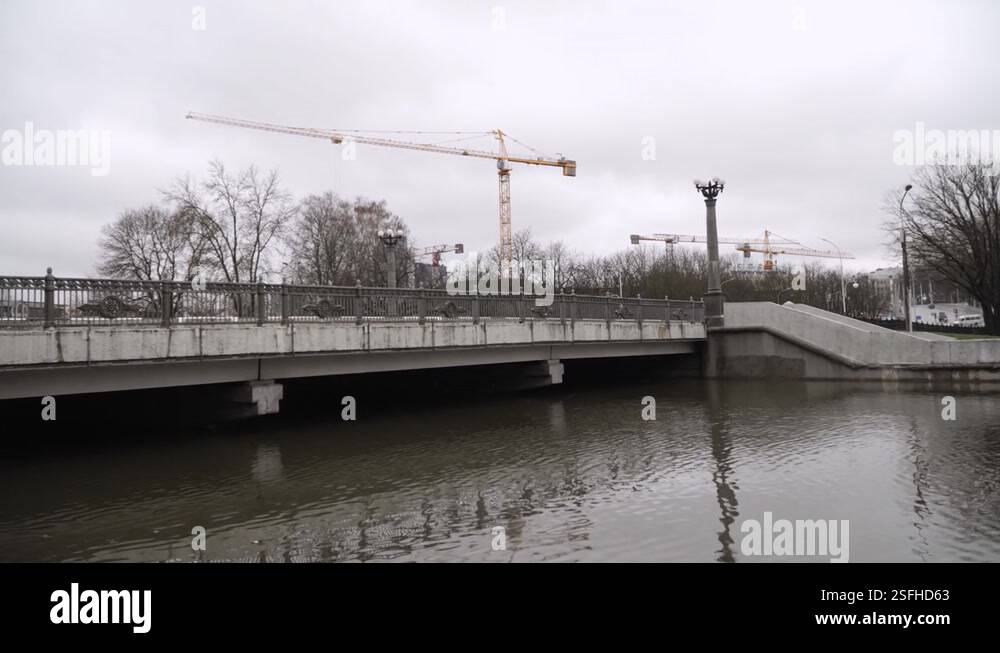 Concrete wet bridge across city river, trees and building cranes on ...