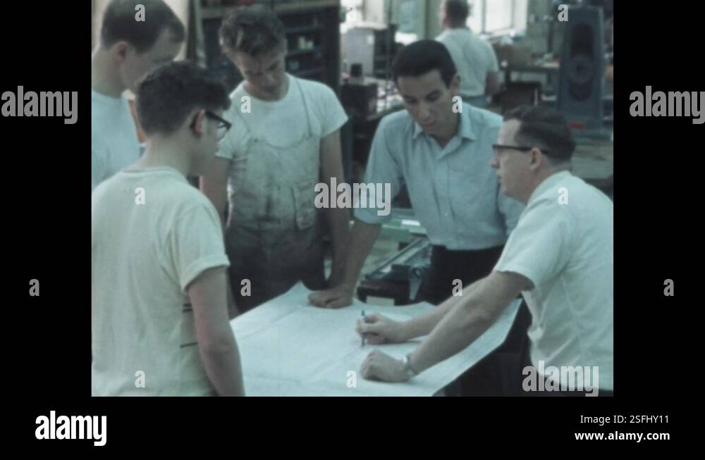 1960s: staff gather around technical drawings in workshop. Man explains ...