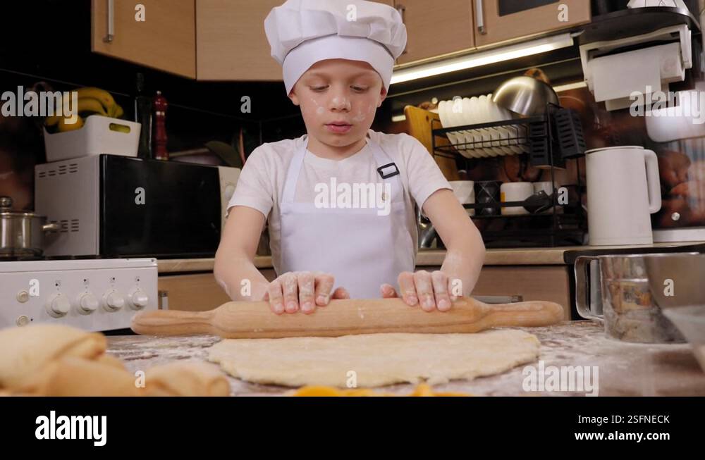 A little boy in an apron rolls out the dough using a rolling pin in the ...