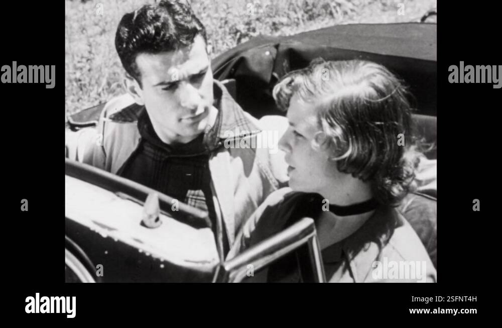 1950s: Teen girl and boy sit in convertible car with her in the driver ...