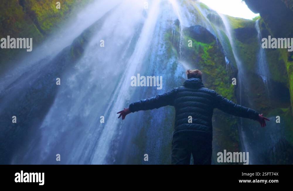 Man standing under Gljufrabui Waterfall with his hands lifted up ...
