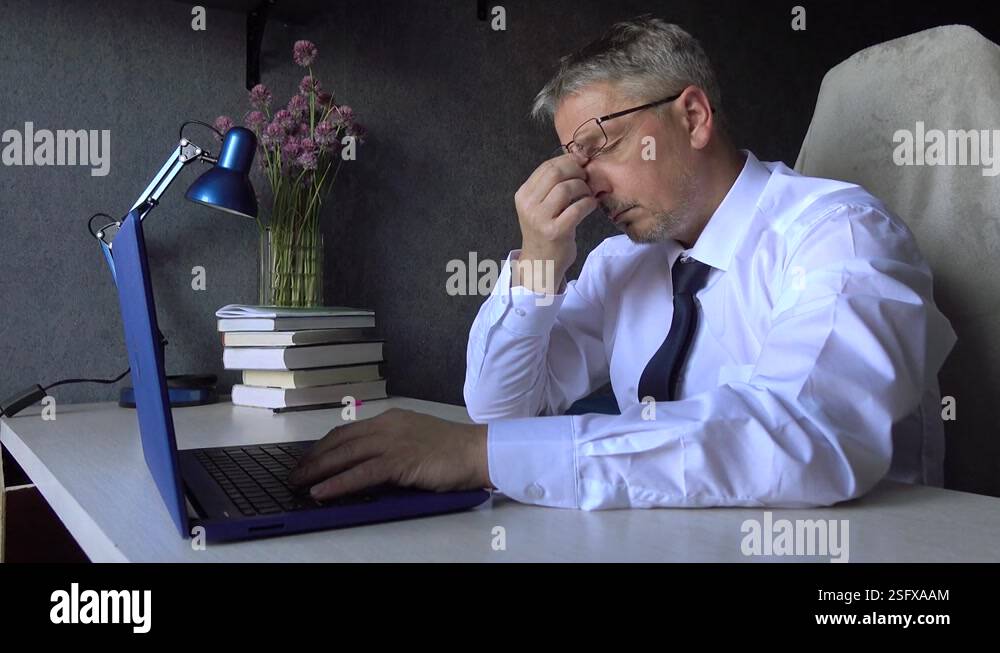A tired man in a white shirt is working at home on a laptop. Remote ...