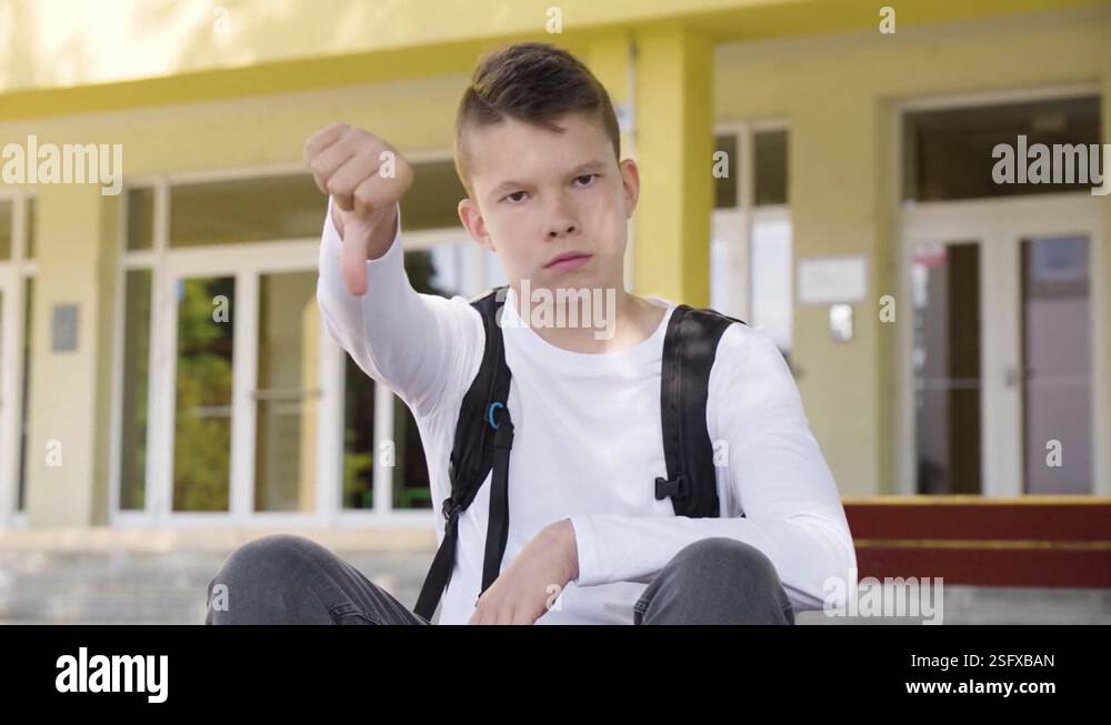 A Caucasian teenage boy shows a thumb down to the camera and shakes his ...