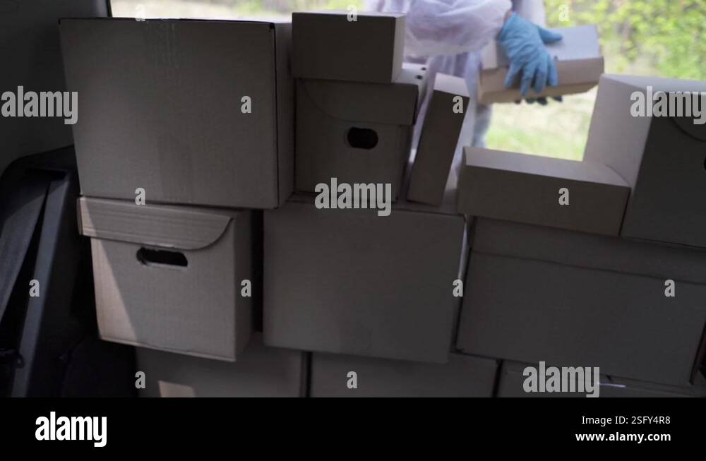 A volunteer in a protective suit loads boxes into the trunk of a car ...