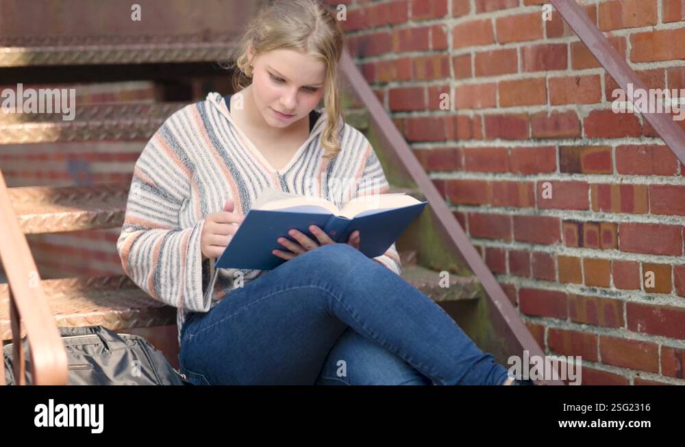 Girl student reading a book outside on the stairs turns a page of her ...