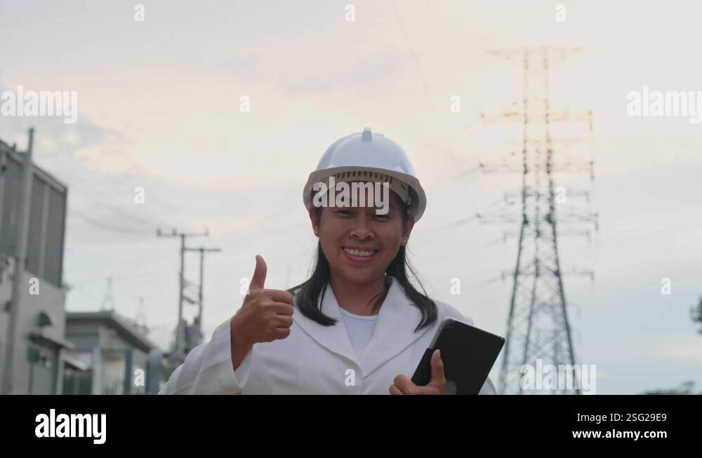 Female electrical engineer thumbs up and smiles on high voltage poles ...