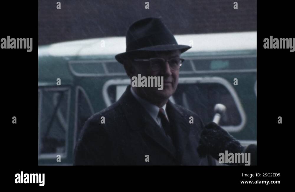 1960s: Man stands outside in snow in suit, hat, speaks into microphone ...
