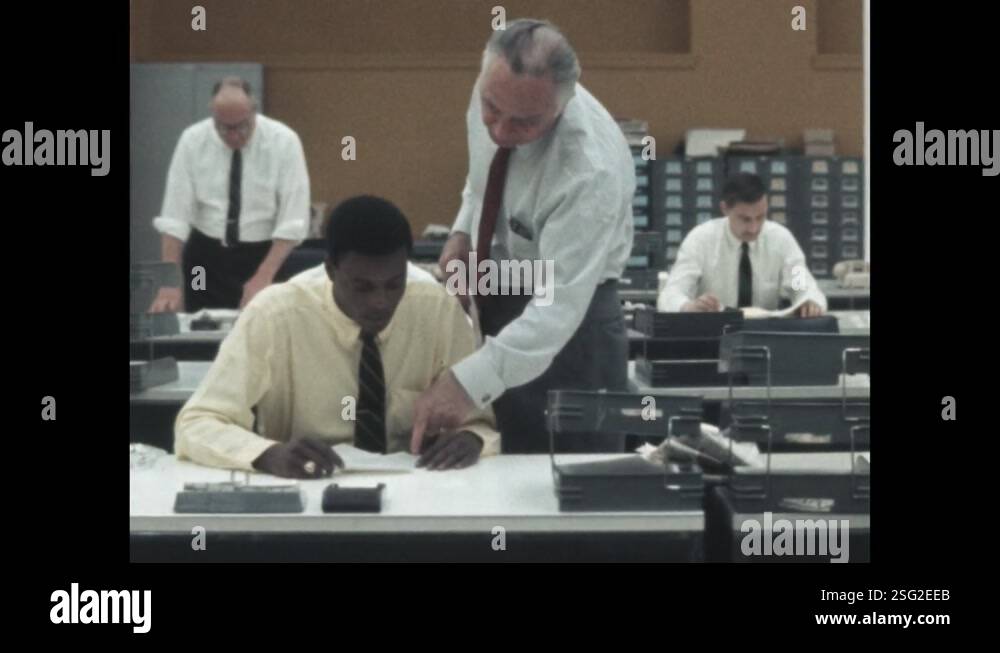 1960s: Open office, people sit at desks, boss leans over man, points at ...