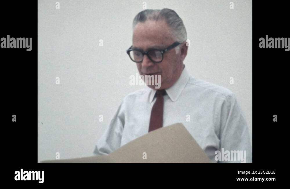1960s: Office, boss stands, holds folder, talks, nods. Man walks past ...