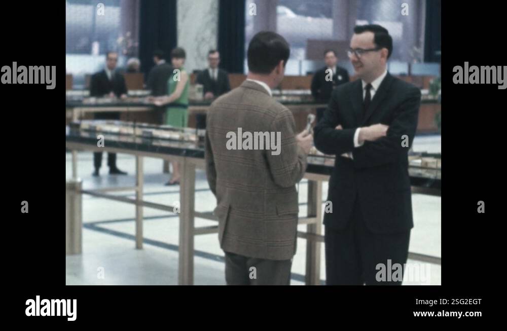 1960s: Bank, man stands, holds microphone, interviews man in suit. Man ...