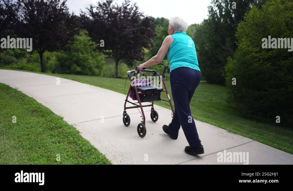 Elderly senior woman pushing a walker, rolling it on a sidewalk outside ...