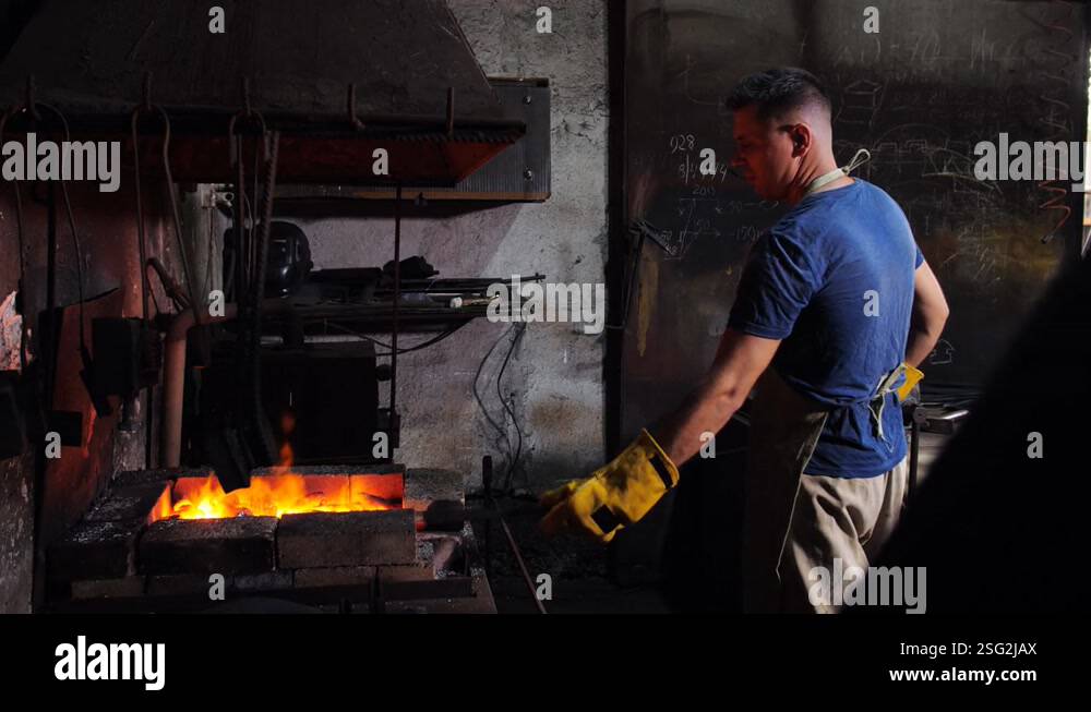 A young blacksmith is stirring the coals with a poker in the forge ...