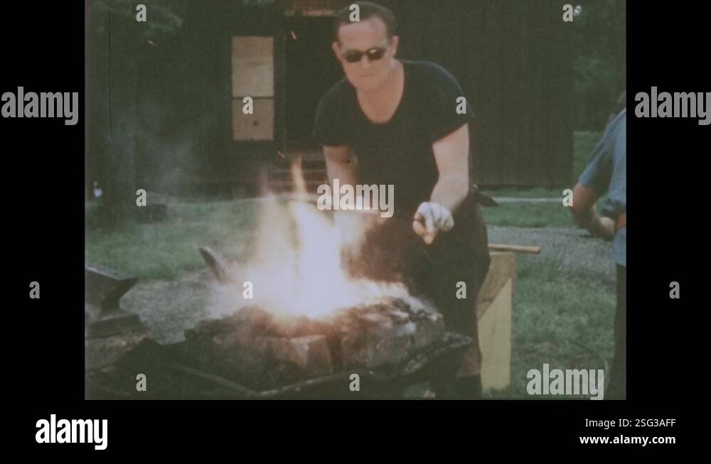 1970s: flames in fire pit. Blacksmith turns hand bellows. Blacksmith ...