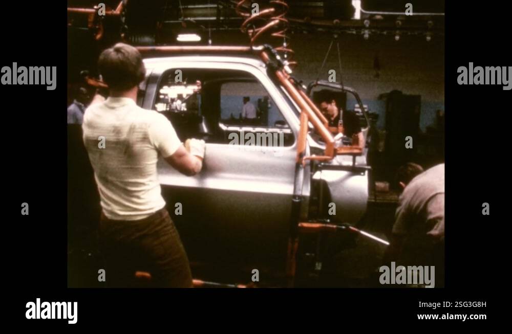 1970s: Workers assembling car body in auto plant. Men lowering engine ...