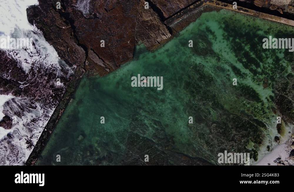 Man swims in rocky tidal pool on Hermanus coastline; drone top down ...