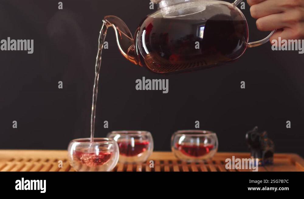 Lifting glass teapot, pouring hot black tea into a glass cup standing ...