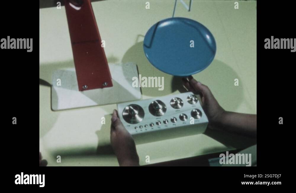1970s: Boy talking. Hands slide box onto table, point to weights. Close ...
