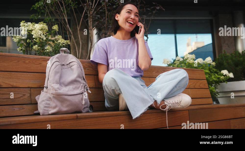 Asian woman sitting on bench talks on phone with friend Stock Video Footage - Alamy