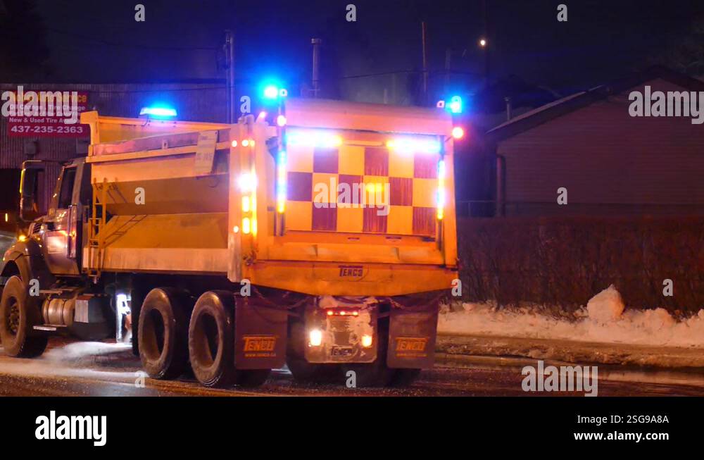 A snow plow disperses salt at night while reversing down an ice-covered ...