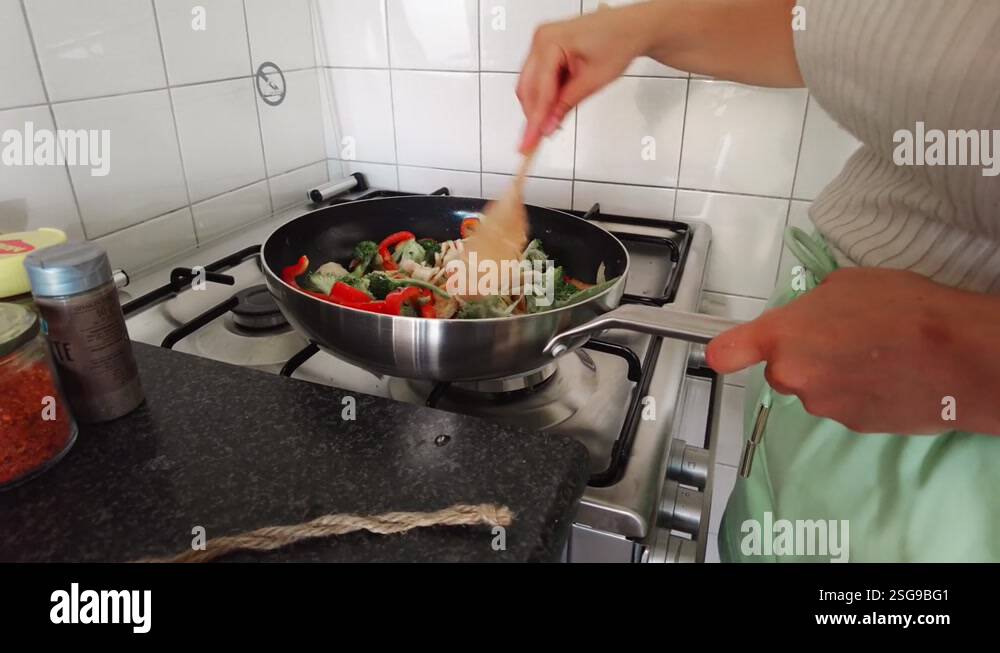 woman stirs wooden spoon in frying pan full with vegetables broccoli ...