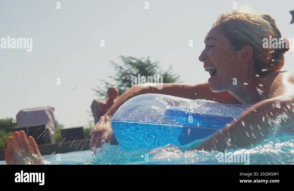 Couple With Man Splashing Woman Floating On Airbed In Swimming Pool On ...
