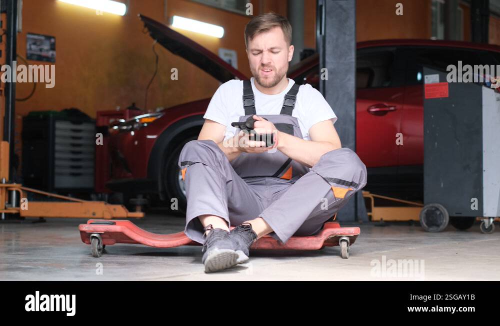 A very angry young car mechanic sitting in his workshop takes off his ...