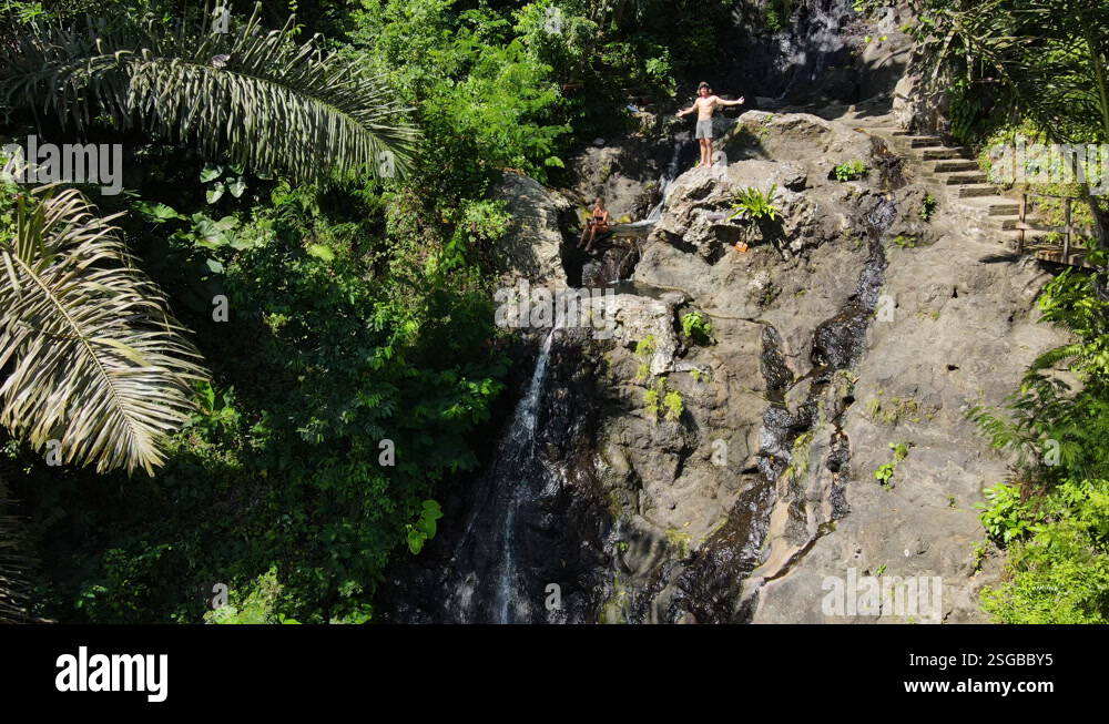 She and he sit on rocks on the top of an exotic waterfall surrounded by ...