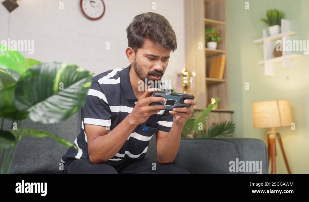 young man playing video game on mobile phone while sitting on sofa at ...