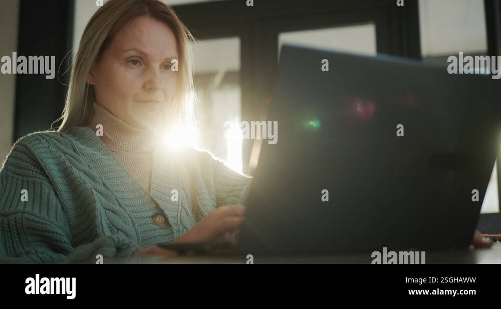 Middle-aged woman using a laptop, the sun from the window illuminates ...